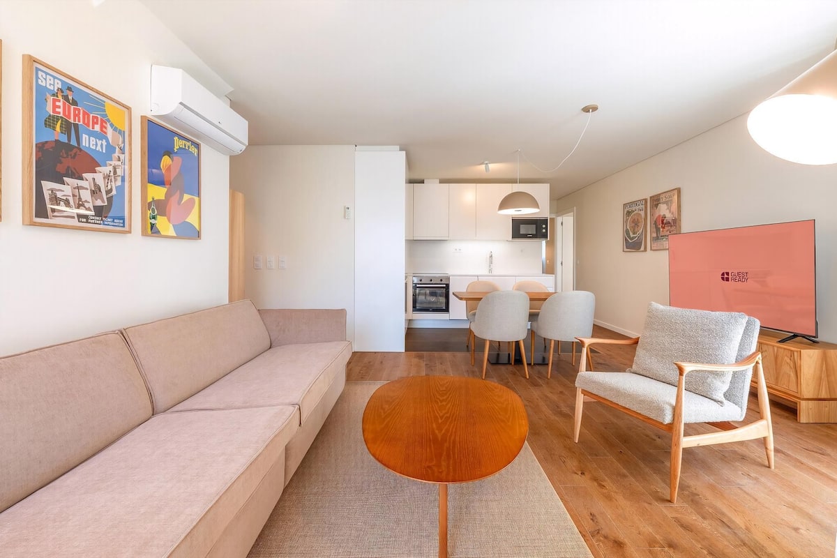 Uma sala de estar com sofá, mesa de centro redonda de madeira e poltrona cinza. Há uma área de jantar com mesa e cadeiras, e uma cozinha com micro-ondas e forno.