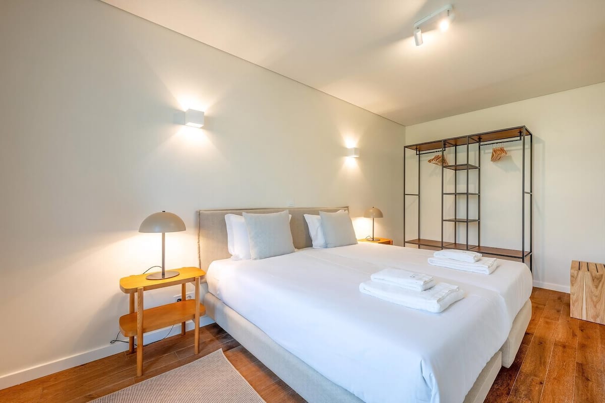 Quarto com cama grande, abajures, mesa de cabeceira de madeira, espelho e um banco de madeira.