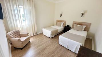 Quarto de hotel com duas camas, uma cadeira, uma escrivaninha e um abajur.