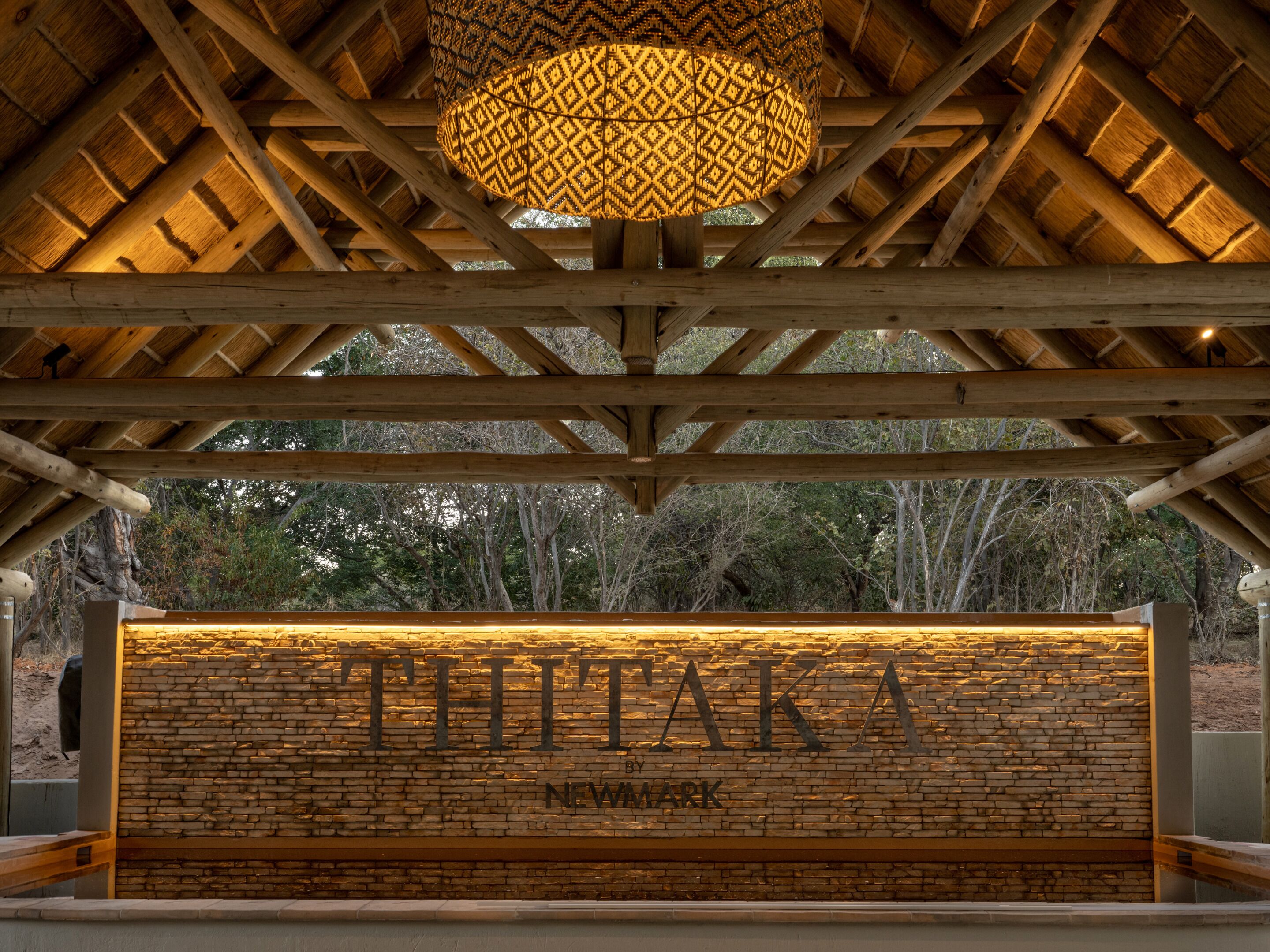 Estrutura de madeira com luminária pendente e parede de pedra com os nomes 'THITAKA' e 'NEWMARK' gravados.