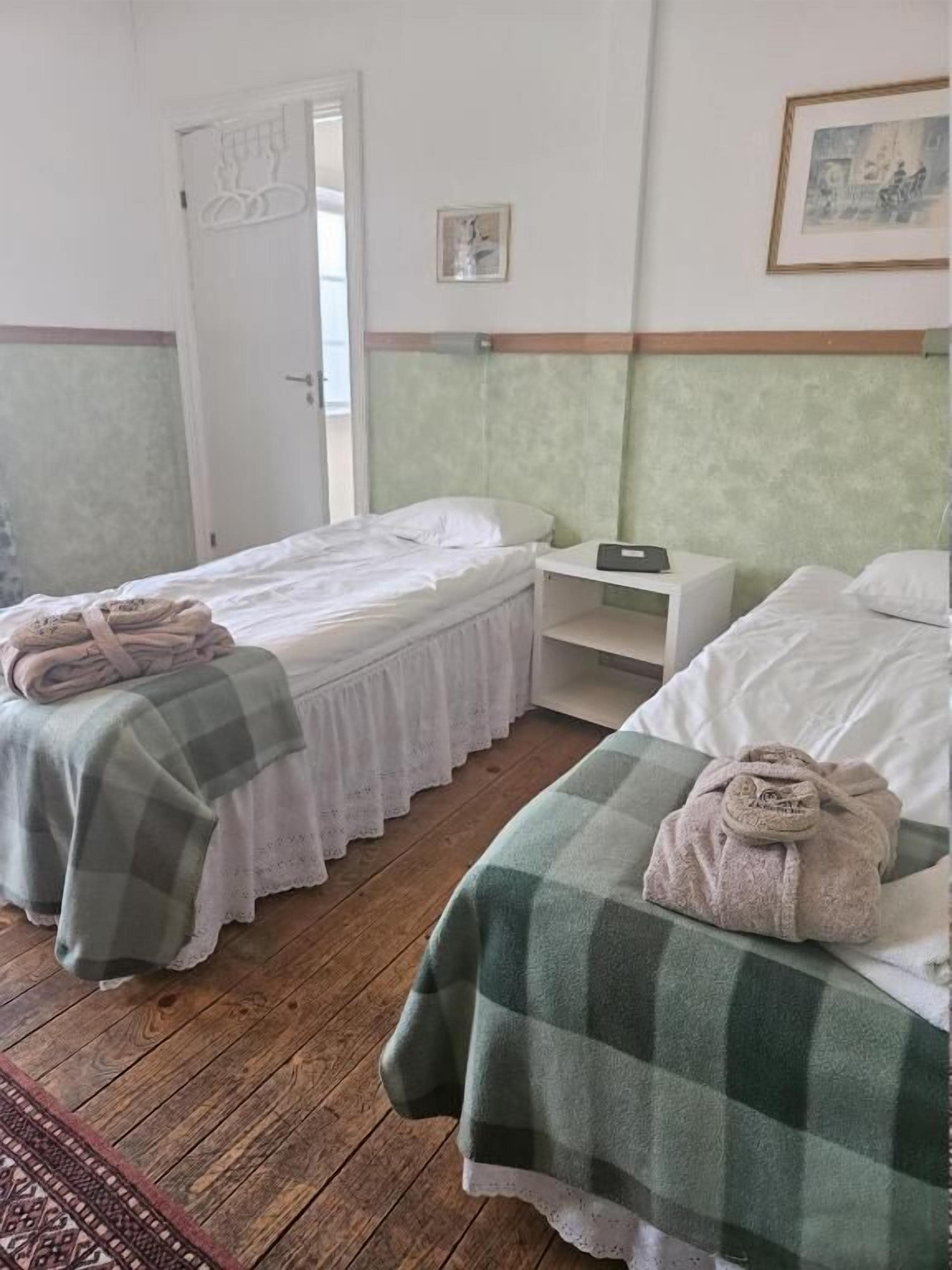 Quarto com duas camas, cada uma com colcha branca e manta verde xadrez. Há uma pequena mesa de cabeceira com luminária e um quadro na parede.