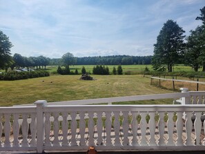Vista de varanda com guarda-corpo branco, sobre um campo gramado com árvores e uma floresta ao longe.