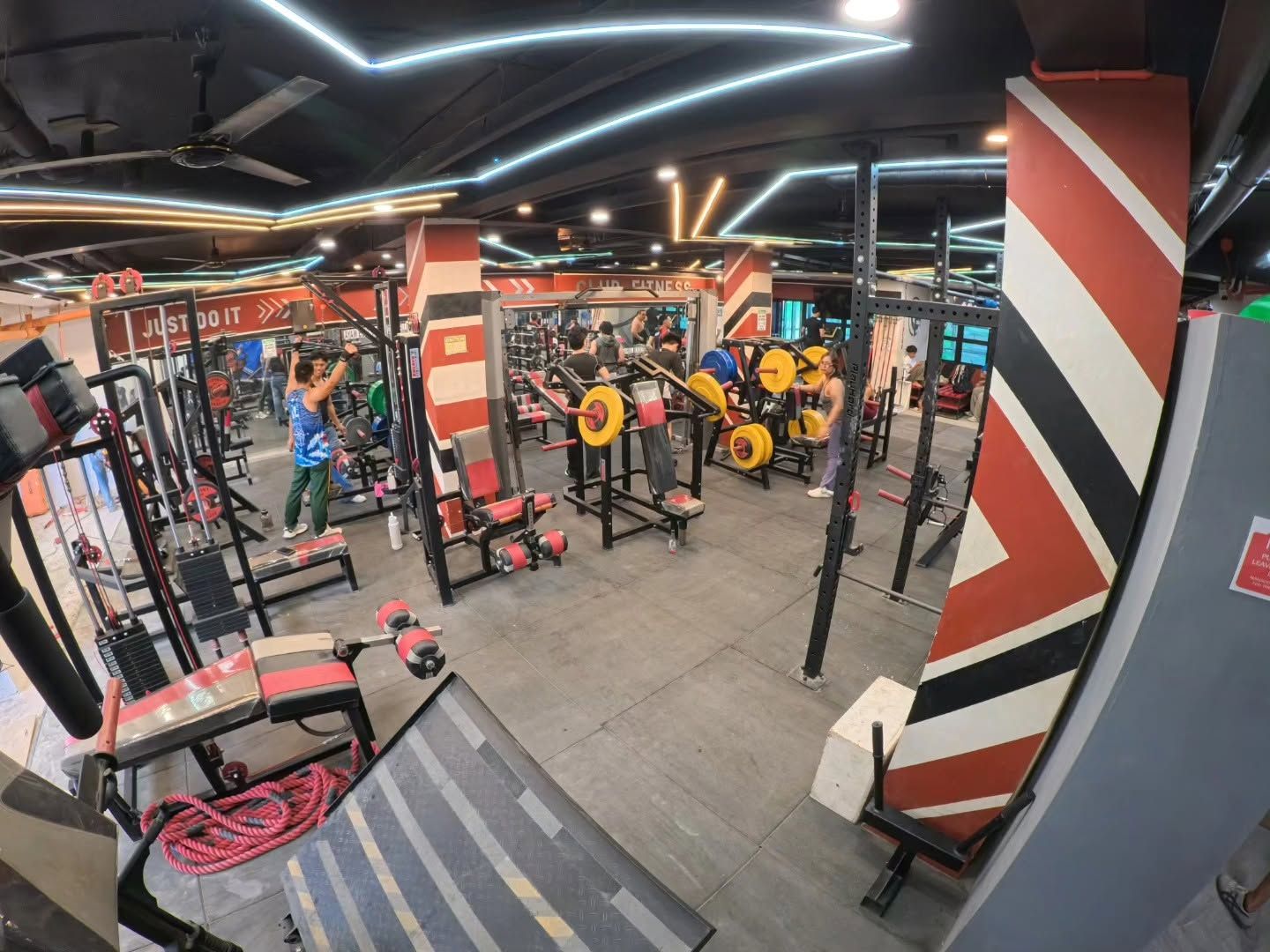 Uma academia com diversos equipamentos e máquinas de musculação, uma parede listrada em vermelho e branco e um teto com luminárias lineares.
