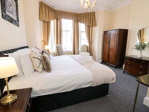 Quarto de hotel com uma cama grande, um guarda-roupa de madeira, uma escrivaninha com abajur e uma janela com cortinas.