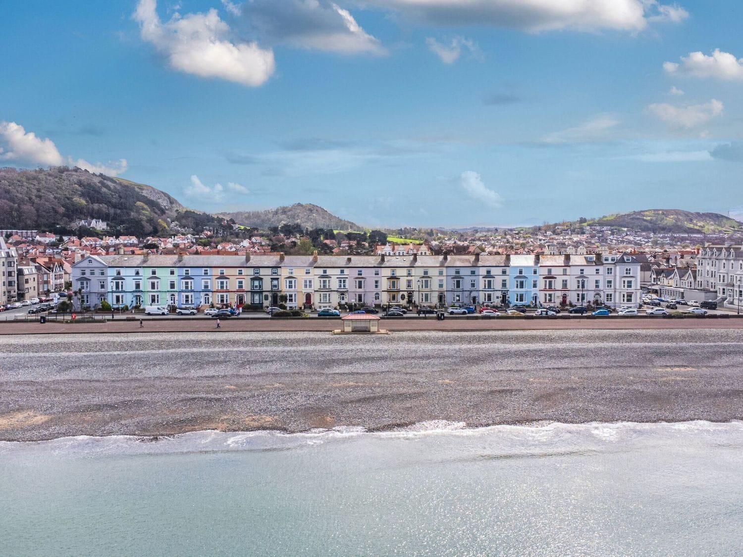 Uma cidade costeira com casas em terraços ao longo da praia, céu claro e colinas ao longe.