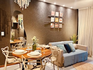 Uma sala de estar moderna com área de jantar, incluindo um sofá, cadeiras, uma mesa de jantar posta para quatro pessoas e uma prateleira com objetos decorativos.
