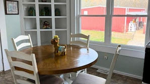 Uma área de refeições com uma mesa redonda de madeira, cadeiras brancas e um cão sobre a mesa. Há uma janela com vista para um edifício vermelho lá fora.