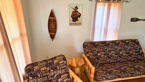 Quarto com dois sofás estampados, uma mesa lateral de madeira, uma prateleira na parede e um cartaz.
