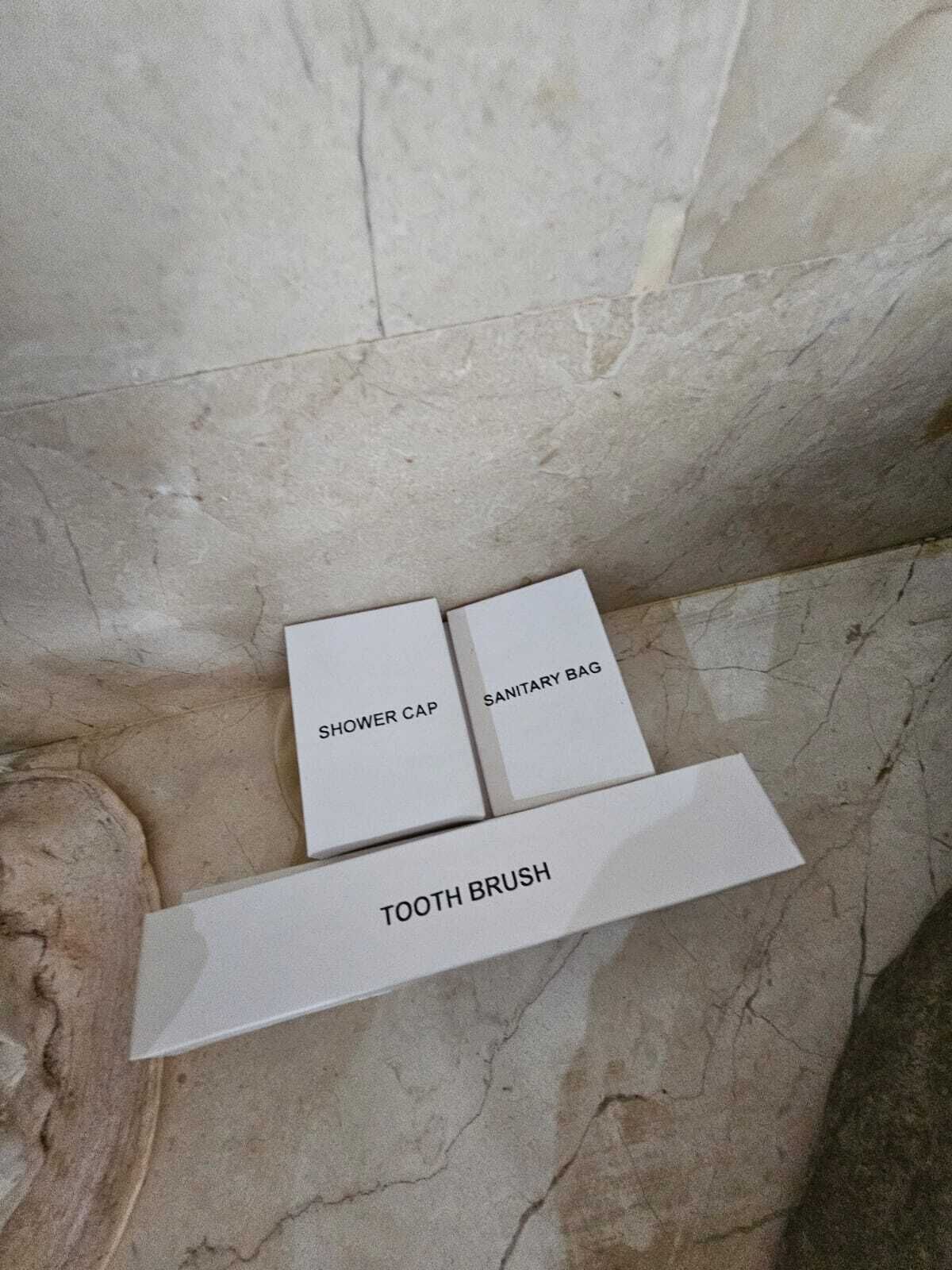 Três caixas de higiene pessoal identificadas com 'SHOWER CAP', 'SANITARY BAG' e 'ESCOVA DE DENTES' sobre um chão de mármore.