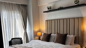Quarto de hotel moderno com uma cama grande, uma poltrona, mesa de cabeceira, abajur e prateleira com uma planta.