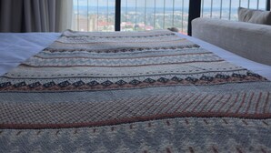 Quarto de hotel com cama, vista da cidade através de grandes janelas e varanda com guarda-corpo.