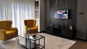 Uma sala de estar moderna com sofá, poltronas, mesa de centro de vidro, TV fixada em uma parede de mármore e um vaso decorativo.
