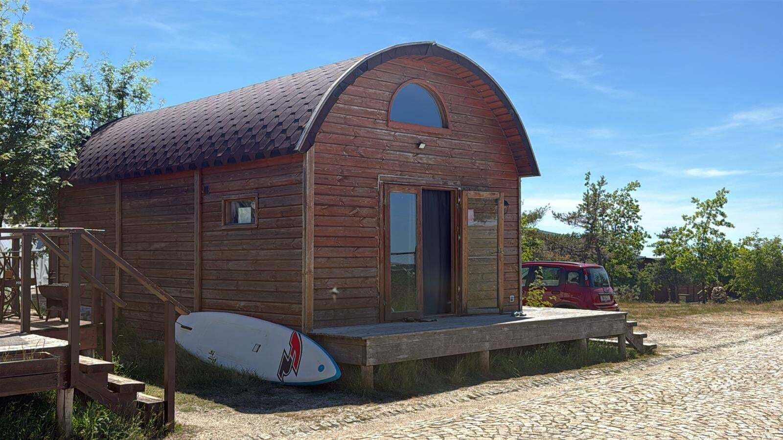 Uma cabana de madeira com telhado inclinado, um pequeno alpendre e uma prancha de surf encostada.