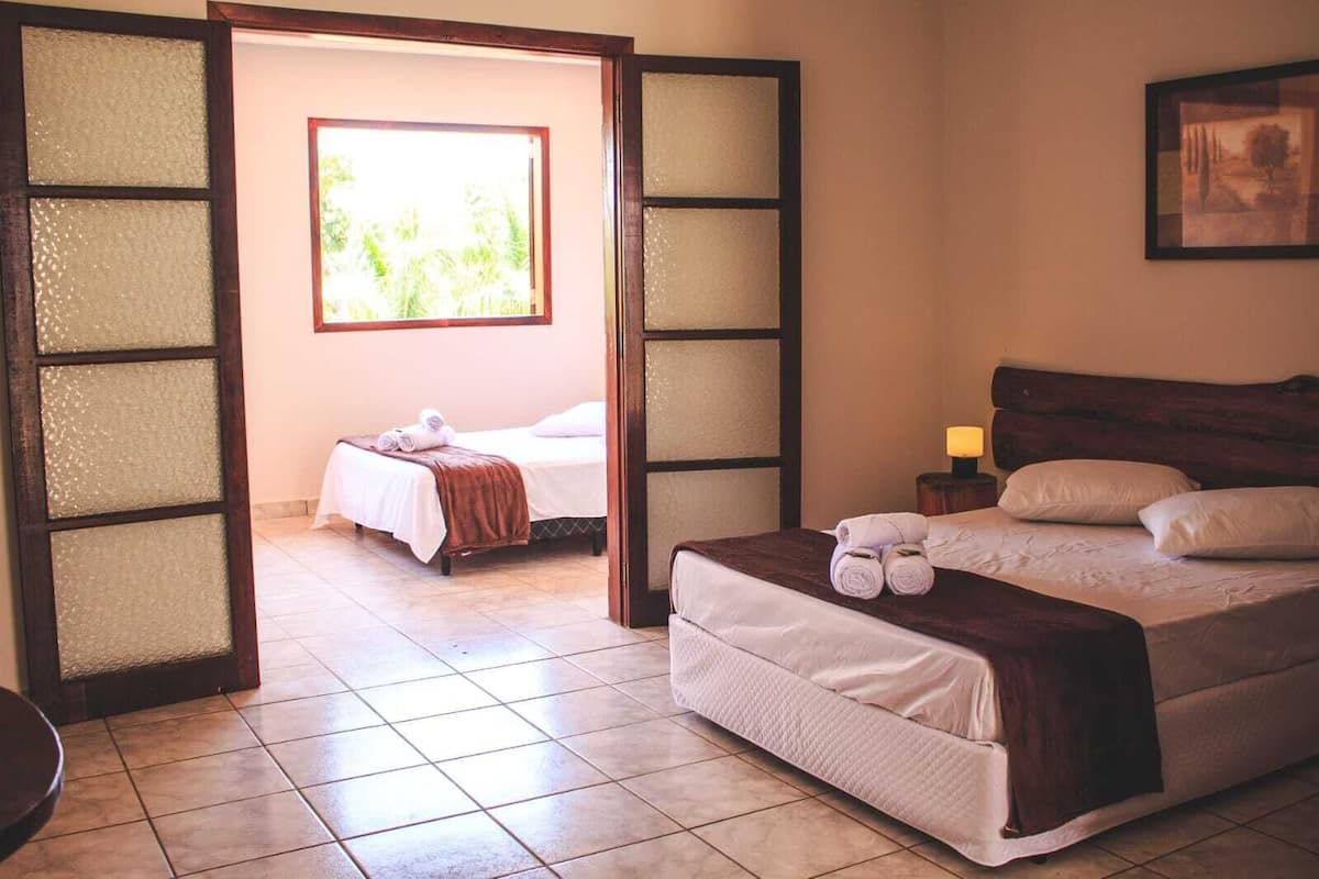 Quarto de hotel com uma cama, mesa de cabeceira e janela com vista para vegetação.