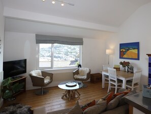 Uma sala de estar com televisão, duas cadeiras, uma mesa redonda, sofá e uma janela com vista, além de um quadro na parede.