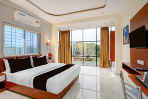 Quarto de hotel com uma cama grande, televisão, secretária e varanda com cortinas.