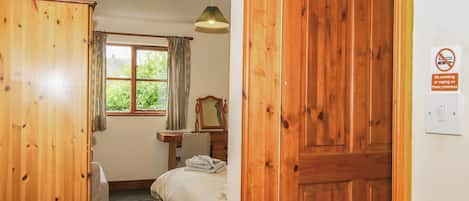 Uma porta de madeira, uma cama, uma escrivaninha, um espelho, uma janela com cortinas e um aviso de 'Proibido fumar'.