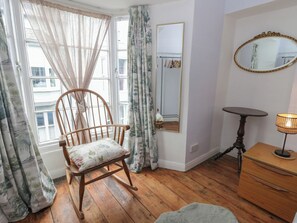 Um quarto com uma cadeira de balanço de madeira, um espelho, uma mesinha com abajur e um penteado.