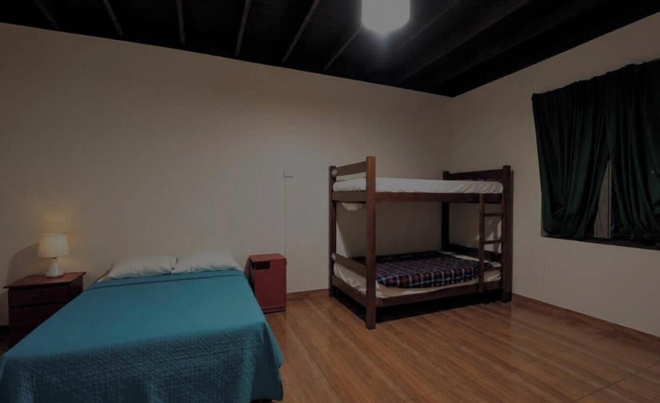 Quarto com cama de solteiro, beliche, mesa de cabeceira, luminária e janela com cortinas.