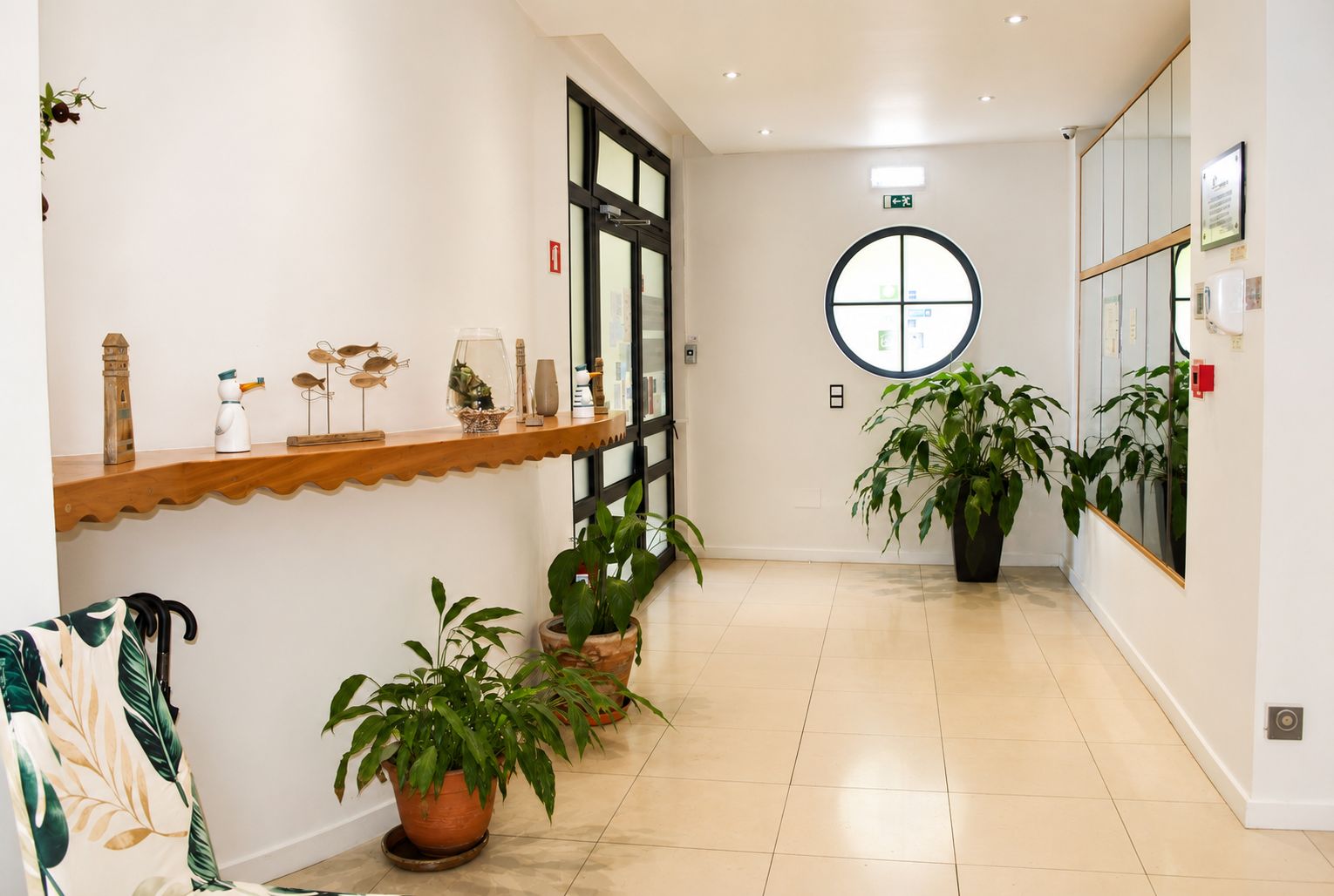 Corredor de hotel com balcão de recepção de madeira, plantas em vasos, janela redonda e porta com alarme de incêndio.
