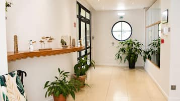 Corredor de hotel com balcão de recepção de madeira, plantas em vasos, janela redonda e porta com alarme de incêndio.