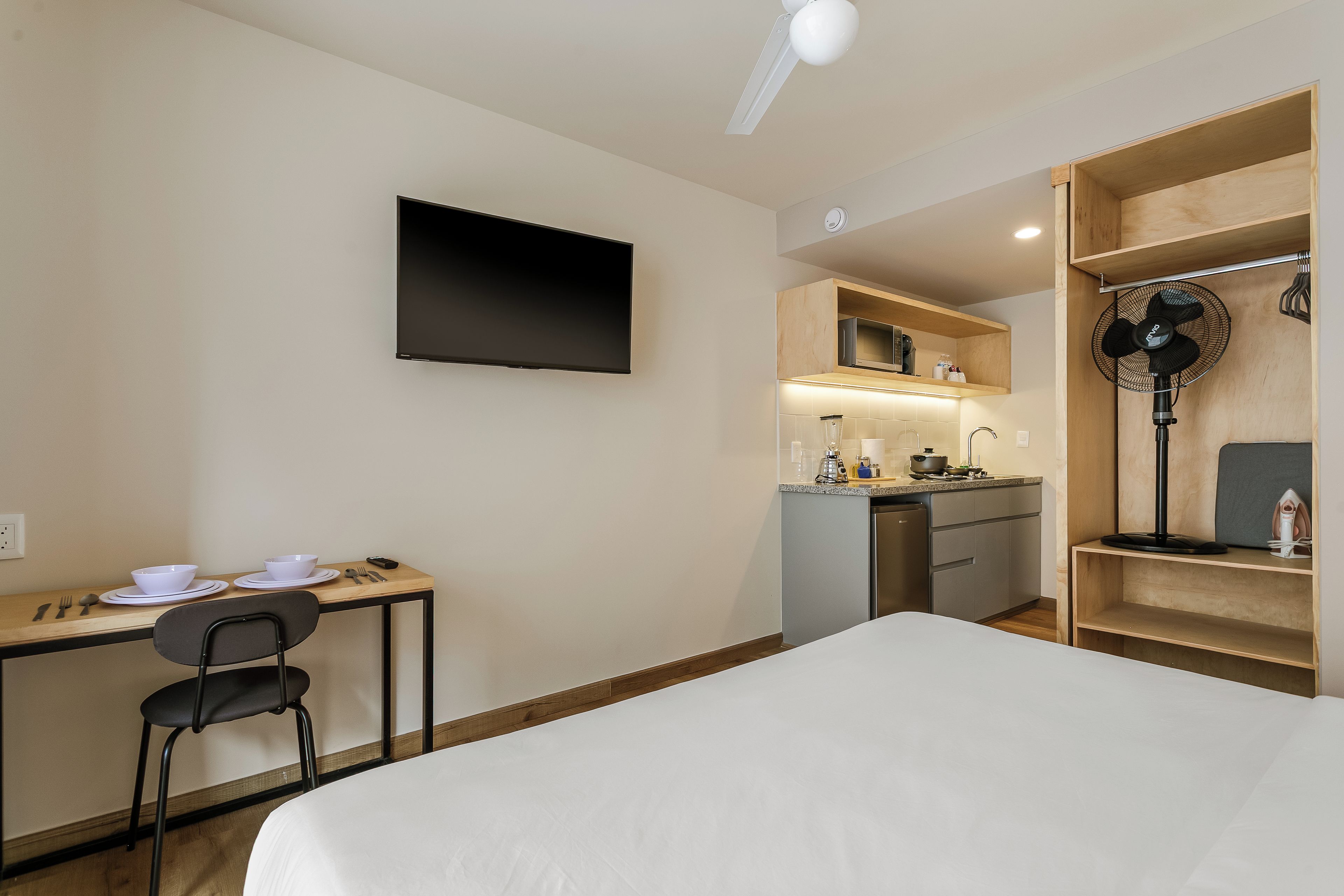Quarto de hotel com cama, escrivaninha e cadeira, televisão, cozinha compacta e ventilador.