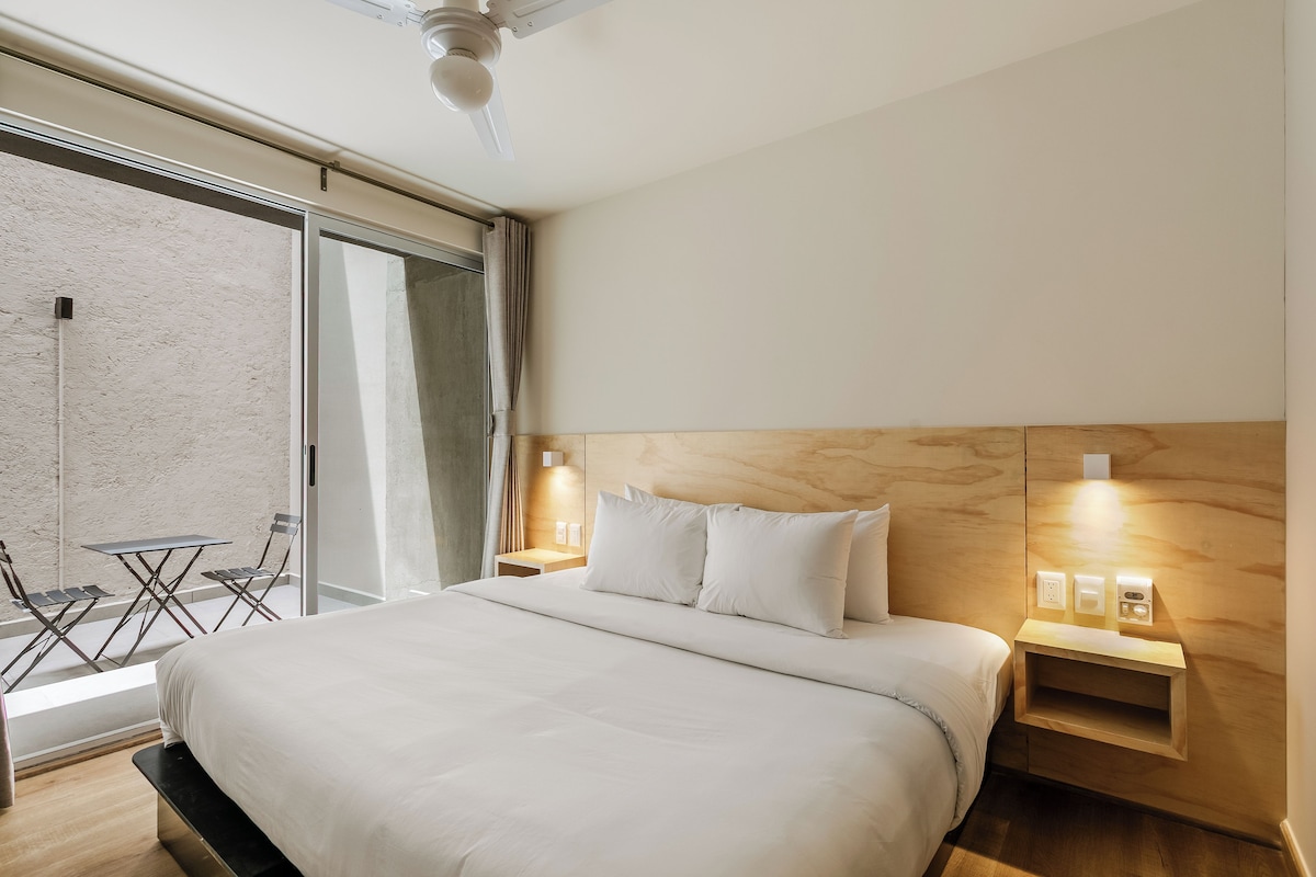 Quarto de hotel com cama, mesa de cabeceira, luminária na parede e vista para o exterior.