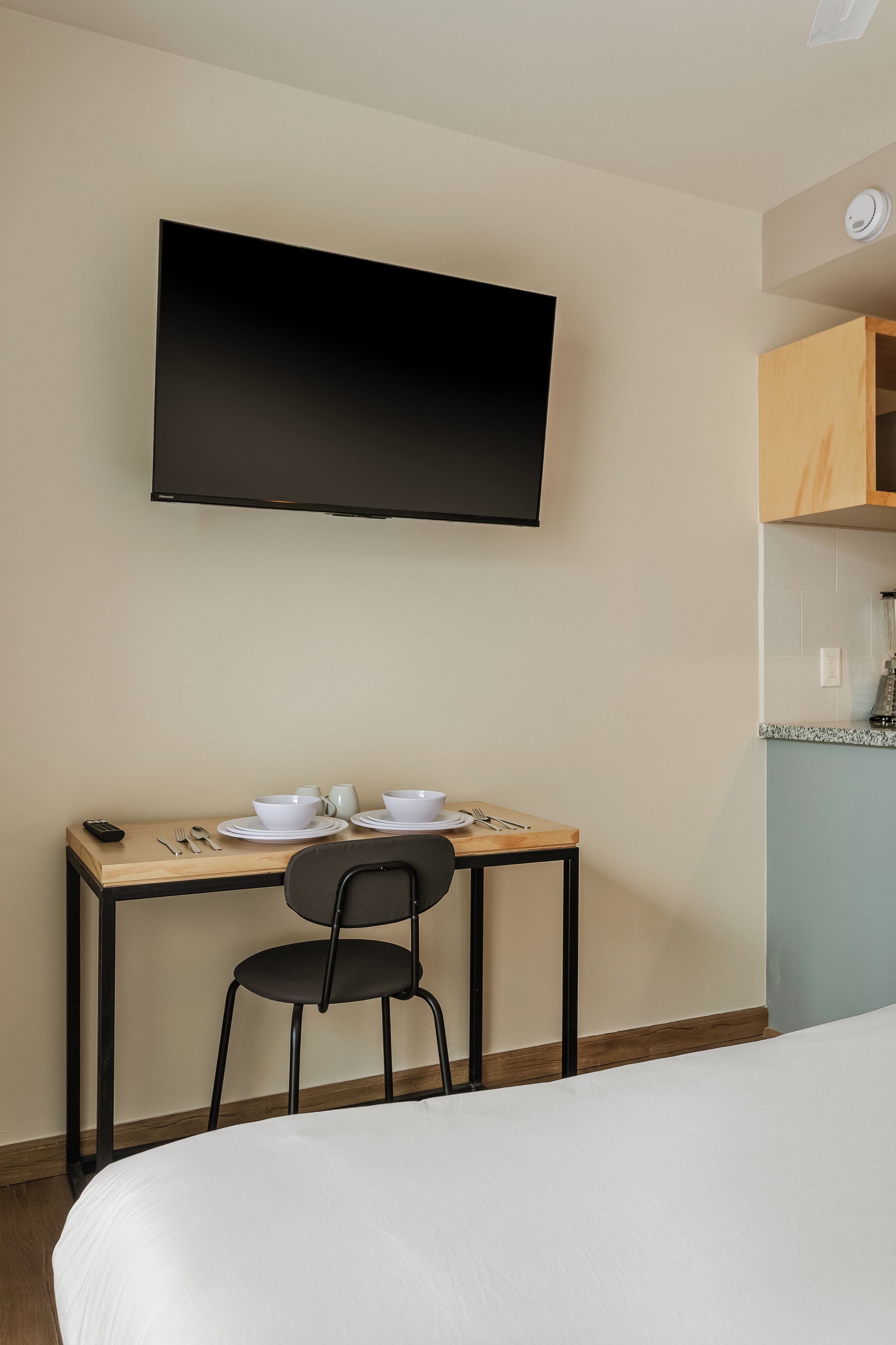 Quarto de hotel com cama, escrivaninha e cadeira, televisão de tela plana na parede e área de cozinha compacta.