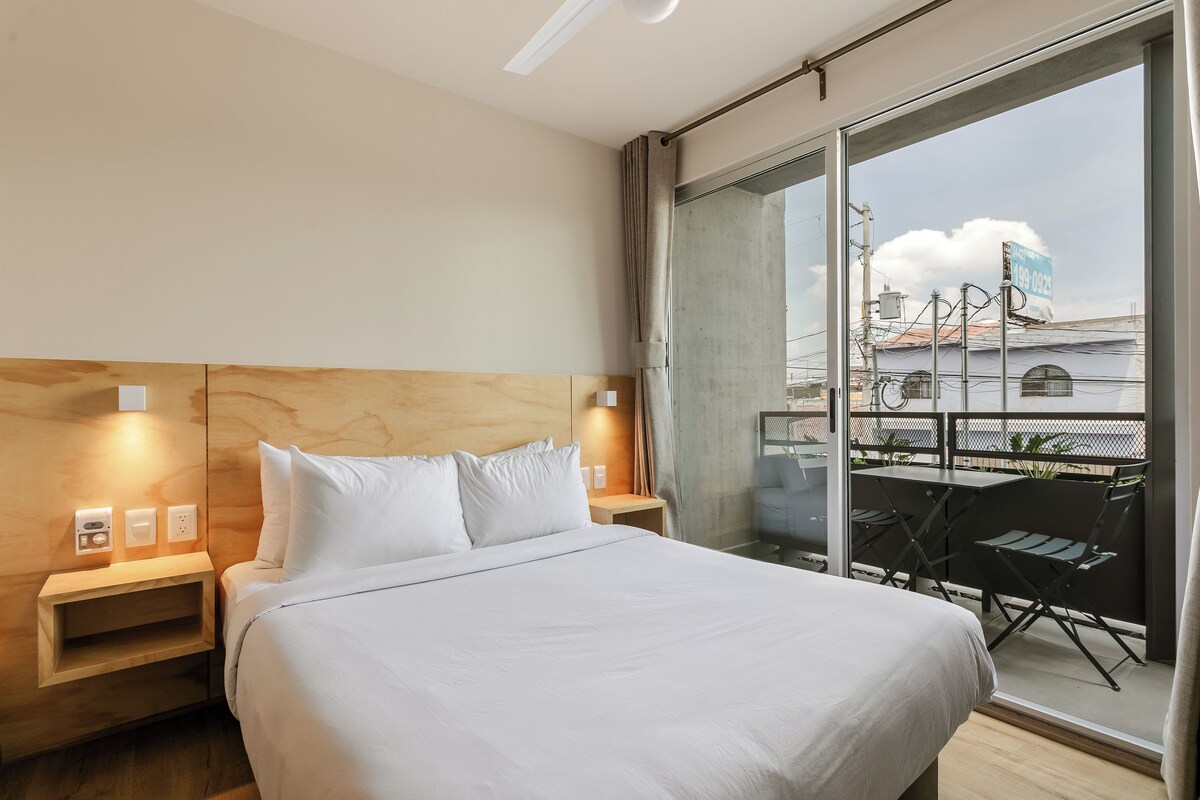 Quarto de hotel com uma cama grande, cabeceira de madeira, mesas de cabeceira e vista da cidade por uma janela ampla.