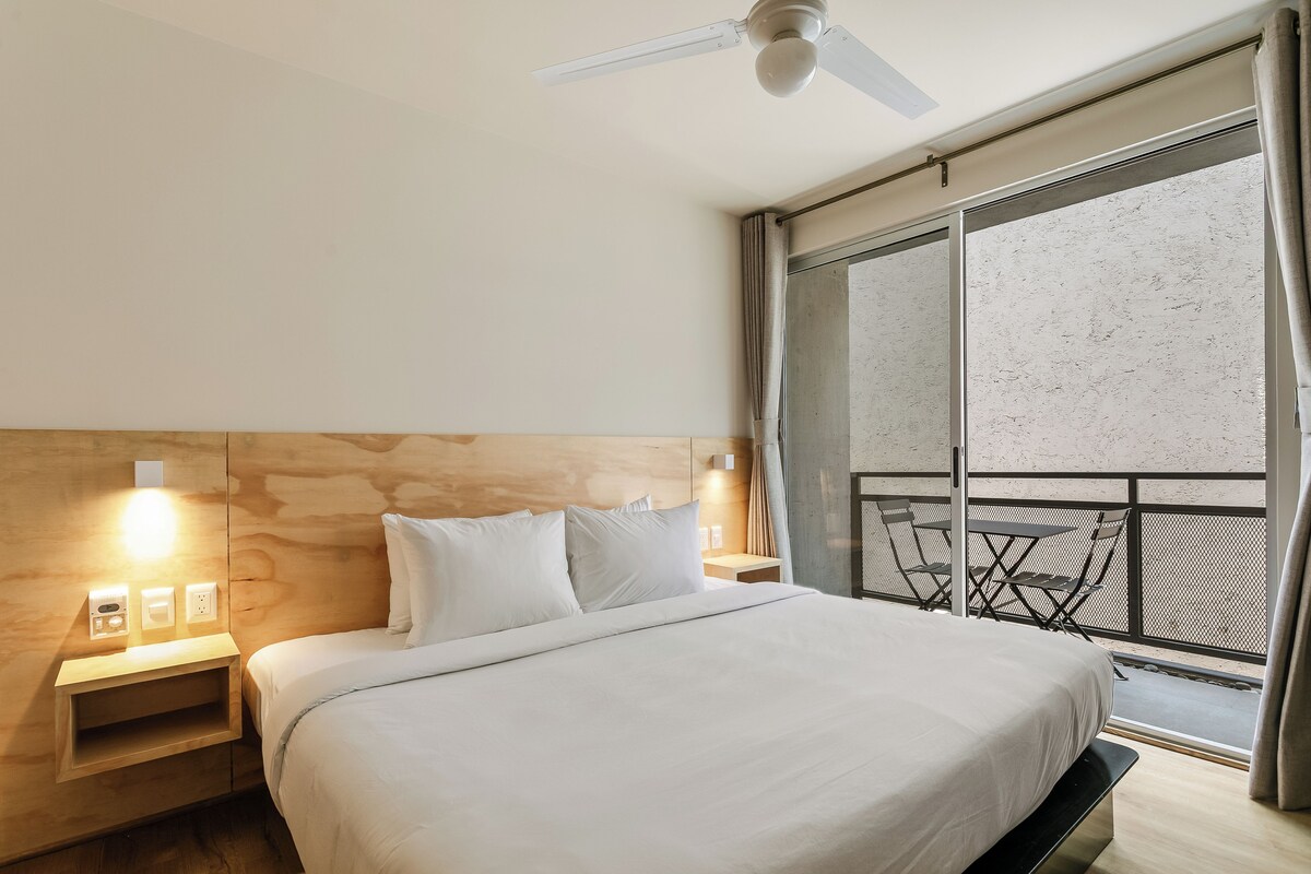 Quarto de hotel com cama, mesa de cabeceira, luminária na parede e vista para o exterior.