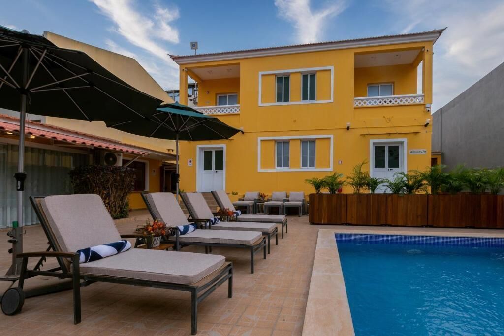 Uma área na piscina com espreguiçadeiras e guarda-sóis, um edifício amarelo com varandas, e um céu azul claro.