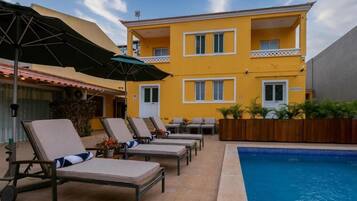 Uma área na piscina com espreguiçadeiras e guarda-sóis, um edifício amarelo com varandas, e um céu azul claro.