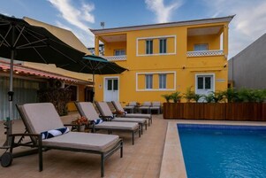 Uma área na piscina com espreguiçadeiras e guarda-sóis, um edifício amarelo com varandas, e um céu azul claro.