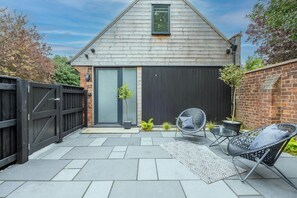 Uma casa com telhado cinzento, porta preta e porta de garagem preta. Há um pequeno jardim com uma planta em vaso e uma cadeira.