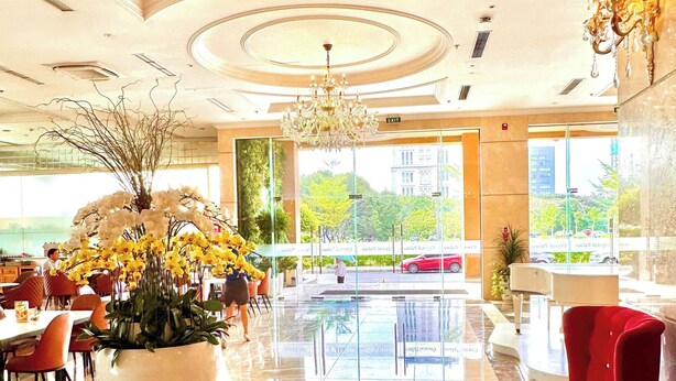 Um saguão de hotel com um lustre grande, uma poltrona vermelha, um vaso com flores amarelas e vista para o exterior através de grandes janelas.