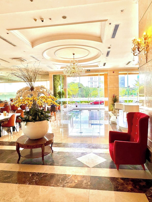 Um saguão de hotel com um lustre grande, uma poltrona vermelha, um vaso com flores amarelas e vista para o exterior através de grandes janelas.