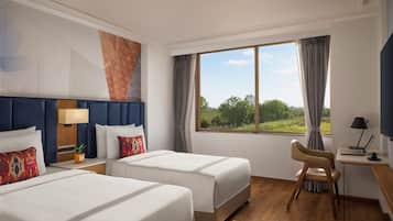 Quarto de hotel com duas camas, uma mesa, uma cadeira, televisão e janela com vista para vegetação.
