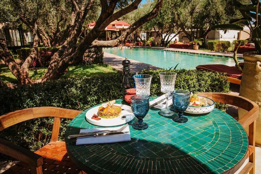 Uma mesa de jantar com pratos de comida, copos e talheres, montada ao ar livre, com uma piscina e vegetação ao fundo.