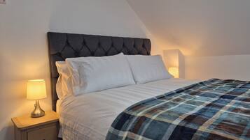 Quarto com cabeceira estofada, cama com roupa de cama branca, manta xadrez, abajur e mesa de cabeceira com gaveta.