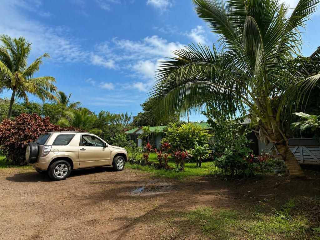 Um SUV bege estacionado em um caminho de terra, com uma casa e vegetação exuberante ao fundo.