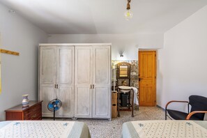 Quarto com armário, mesa de cabeceira, cadeira, cama com colcha estampada, ventilador e banheiro com pia e espelho.