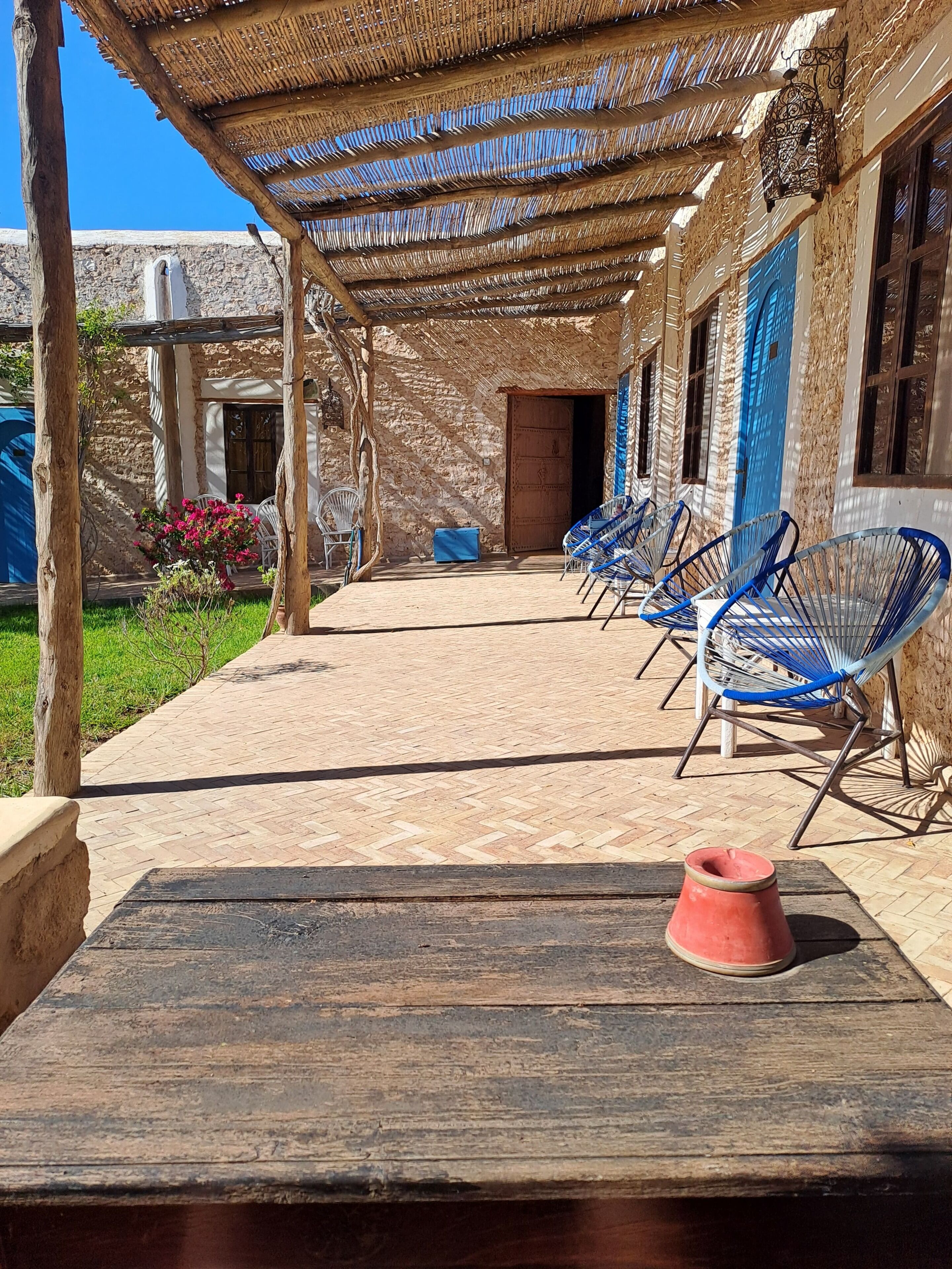 Uma área de estar coberta ao ar livre, com cadeiras e uma mesa, um edifício de pedra com porta, e um céu azul límpido.