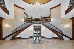 Um hall imponente com teto alto, um lustre, uma escadaria suntuosa com corrimãos ricamente decorados e uma mesa redonda central.