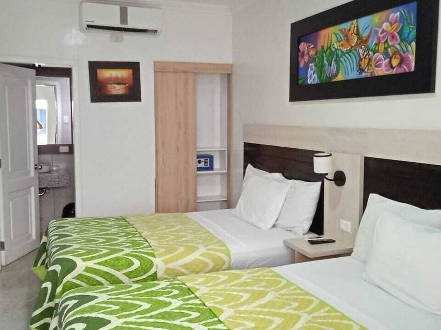 Quarto de hotel com duas camas, uma mesa de cabeceira com abajur, um quadro na parede e um aparelho de ar condicionado.