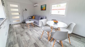 Uma sala de estar moderna com um sofá, uma mesa de jantar com cadeiras, uma mesinha de apoio, um abajur e uma janela com vista para um prédio.