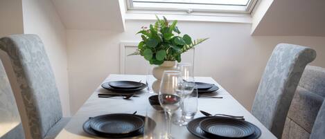 Mesa de jantar posta com pratos pretos, copos de vinho e talheres, com um arranjo de plantas no centro e uma clarabóia acima.