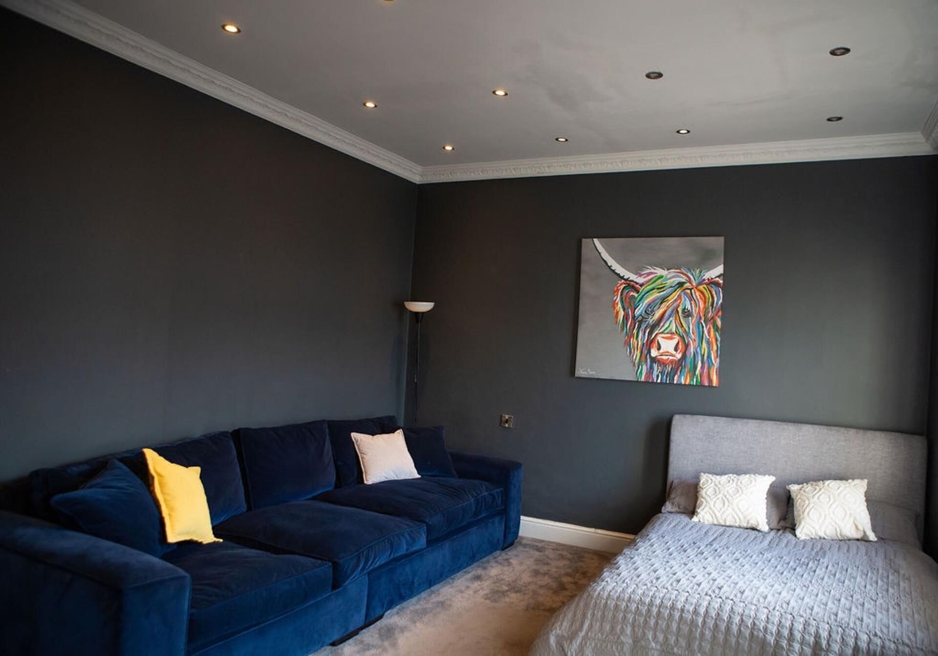 Um quarto com parede cinza escura, um sofá azul com almofada amarela, uma cama com cabeceira cinza e um quadro abstrato colorido.