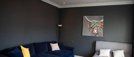 Um quarto com parede cinza escura, um sofá azul com almofada amarela, uma cama com cabeceira cinza e um quadro abstrato colorido.