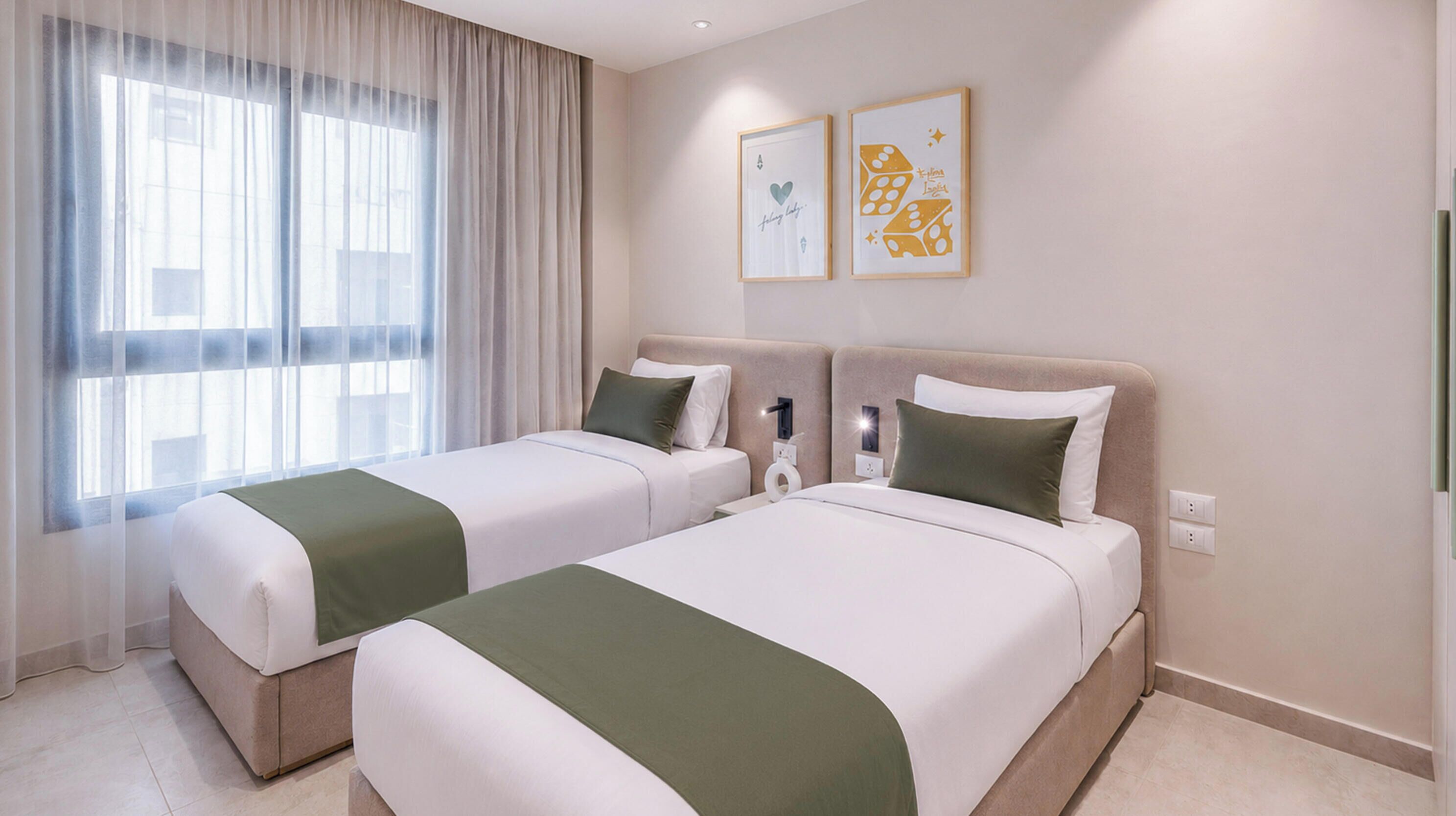 Quarto de hotel com duas camas, uma janela grande com cortinas leves e duas obras de arte nas paredes.