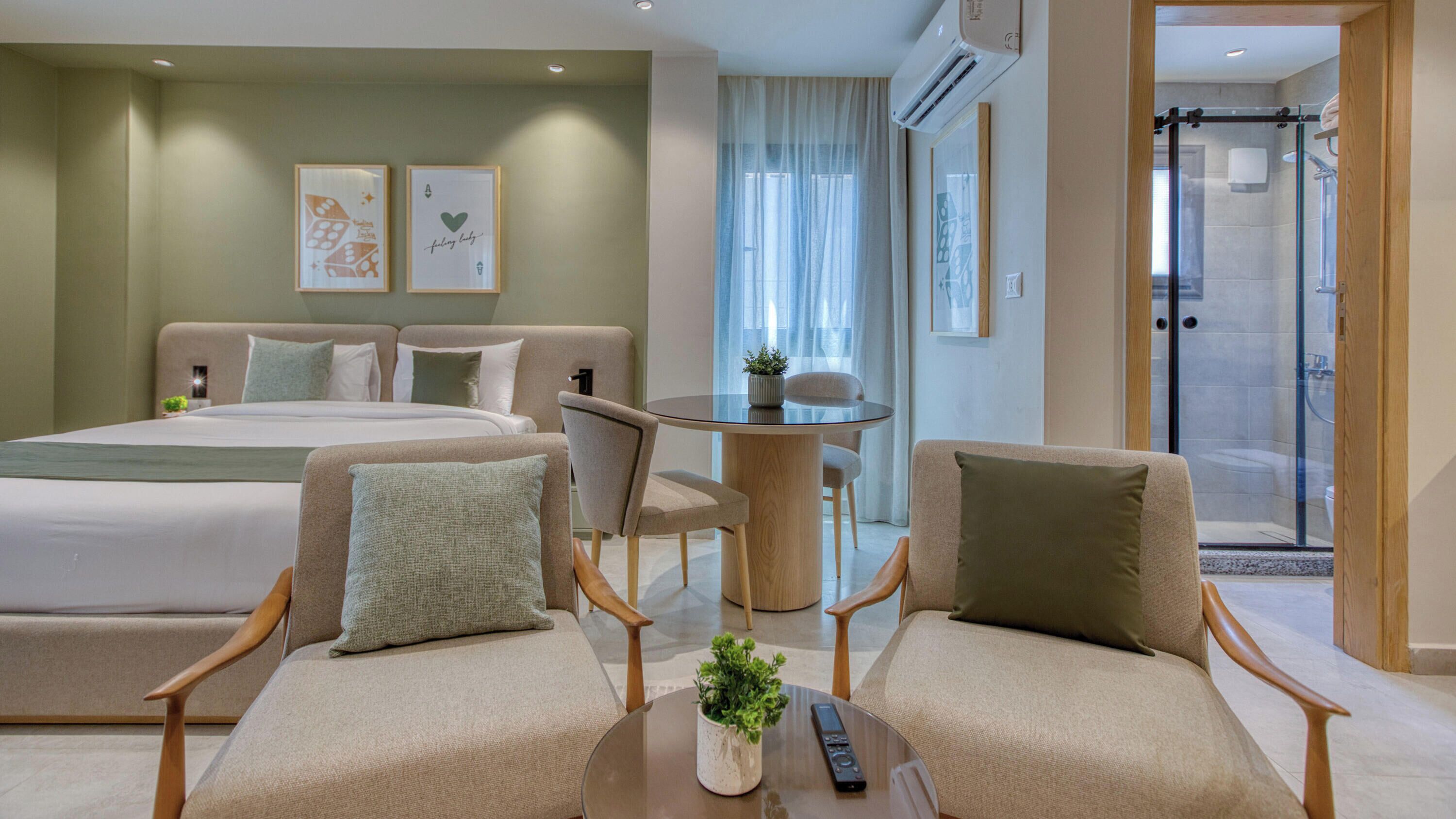 Quarto de hotel com uma cama, duas poltronas, uma mesinha de centro com uma planta e uma área de chuveiro com box de vidro.